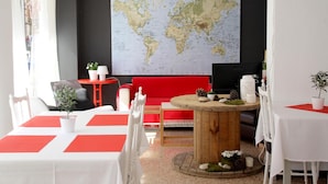 Quarto de hotel com um grande mapa-múndi na parede, um sofá vermelho, uma mesa redonda de madeira com objetos decorativos e mesas brancas com jogos americanos vermelhos.