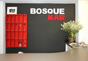 Uma mesa de recepção com um porta-chaves vermelho, uma parede preta com a placa 'BOSQUE MAR' e um vaso com flores brancas.