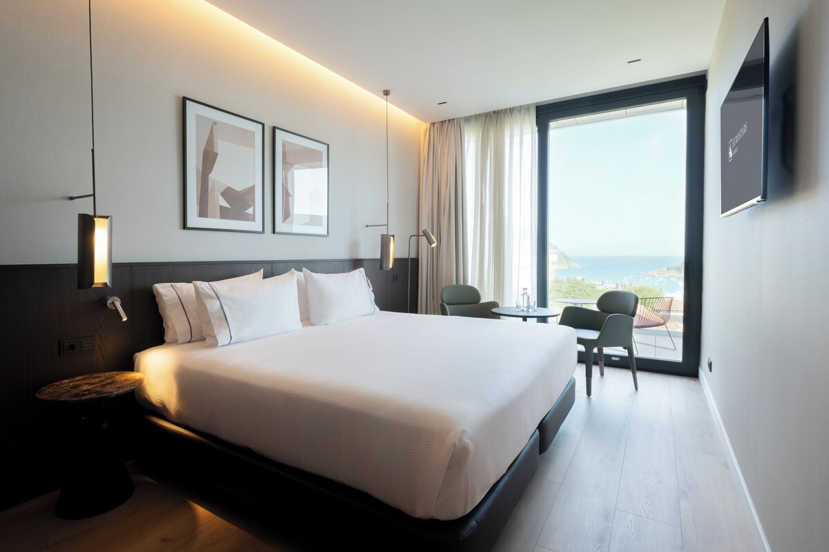 Quarto de hotel com uma cama grande, mesas de cabeceira, uma escrivaninha e vista para o mar.