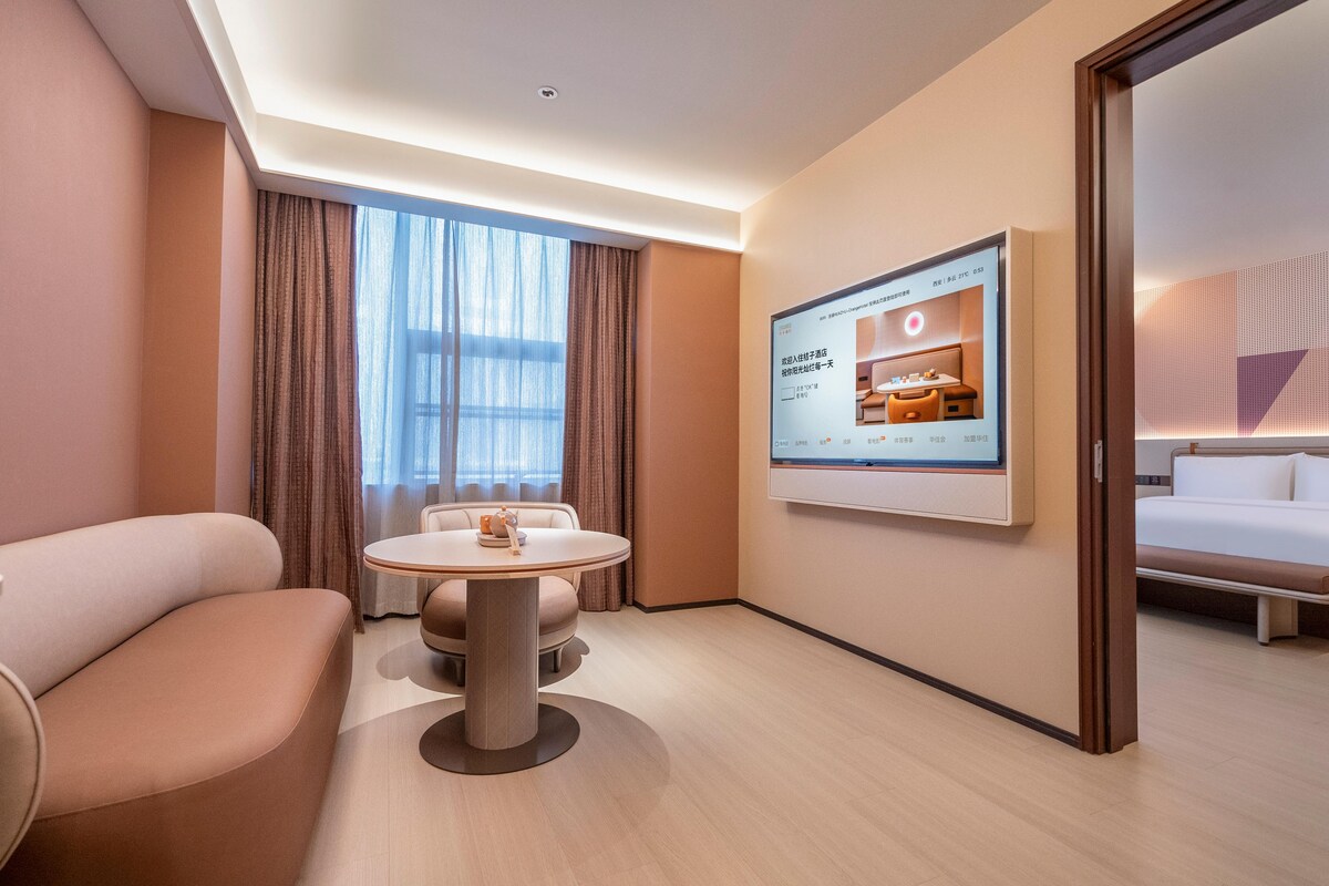 Quarto de hotel moderno com sofá, mesa redonda com bule de chá, cama e televisão na parede exibindo uma imagem de um quarto.