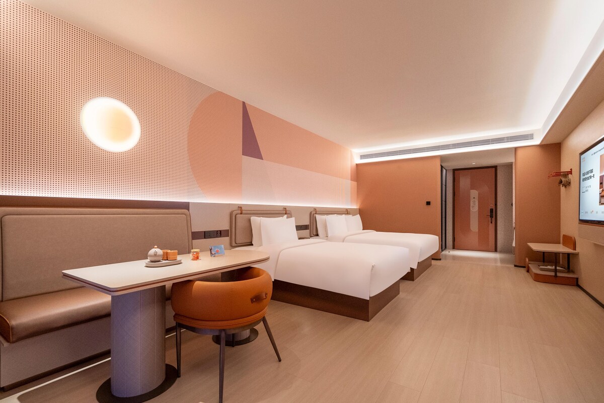 Quarto de hotel moderno com sofá, duas camas de solteiro, uma mesinha com uma cadeira e uma TV grande fixada na parede.