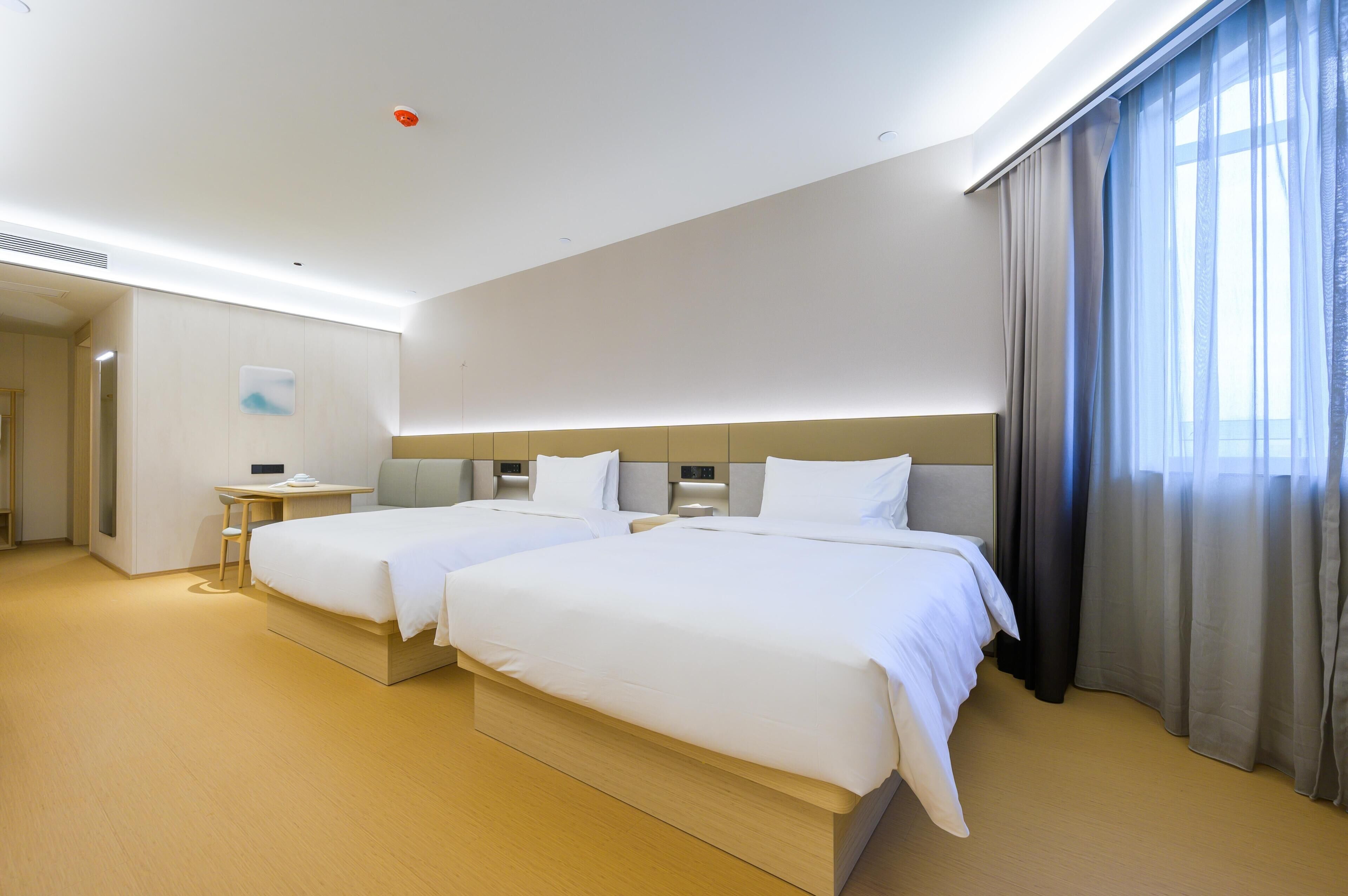 Quarto de hotel com duas camas, uma escrivaninha, uma cadeira e uma janela grande com cortinas.