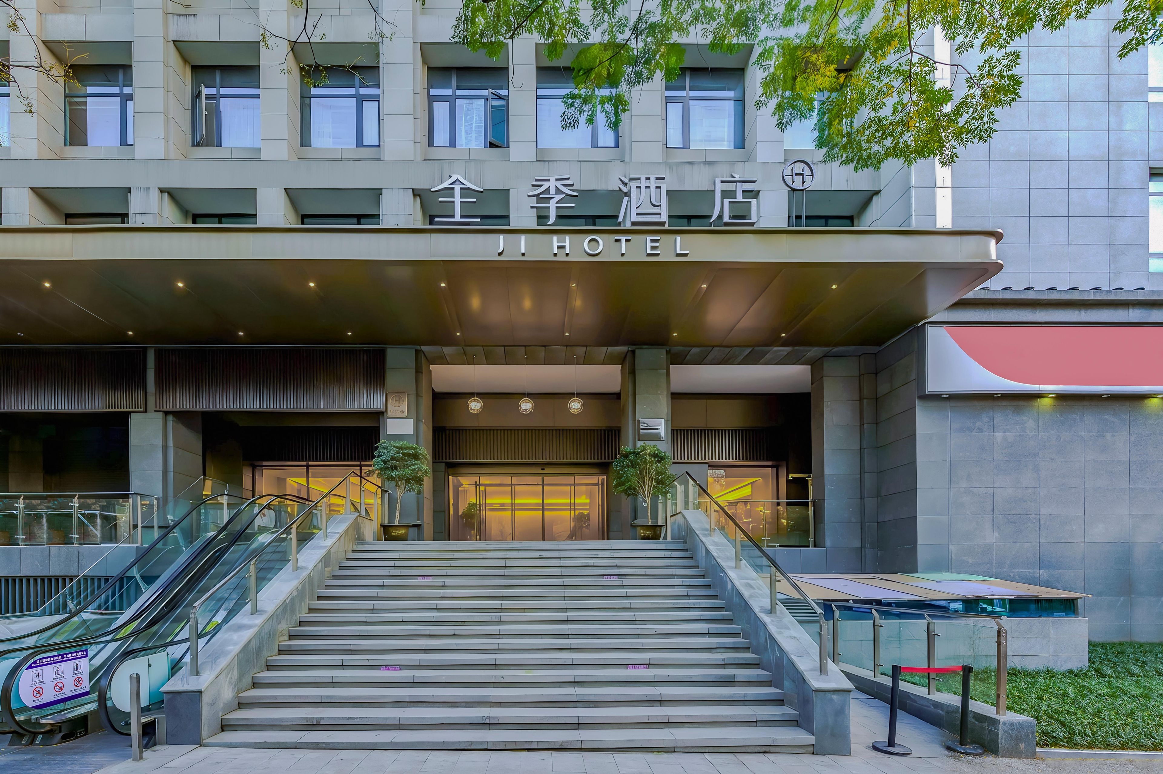 Entrada de hotel com uma escadaria que leva às portas, cercada por guarda-corpos de vidro e plantas em vasos.