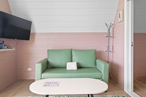 Um quarto com um sofá verde, uma mesa branca, uma televisão presa à parede e um cabideiro.