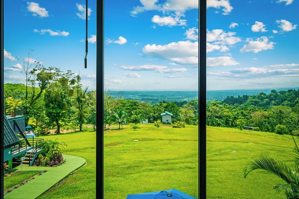 Uma paisagem verdejante com um pequeno edifício branco, céu azul límpido e vista para colinas distantes.