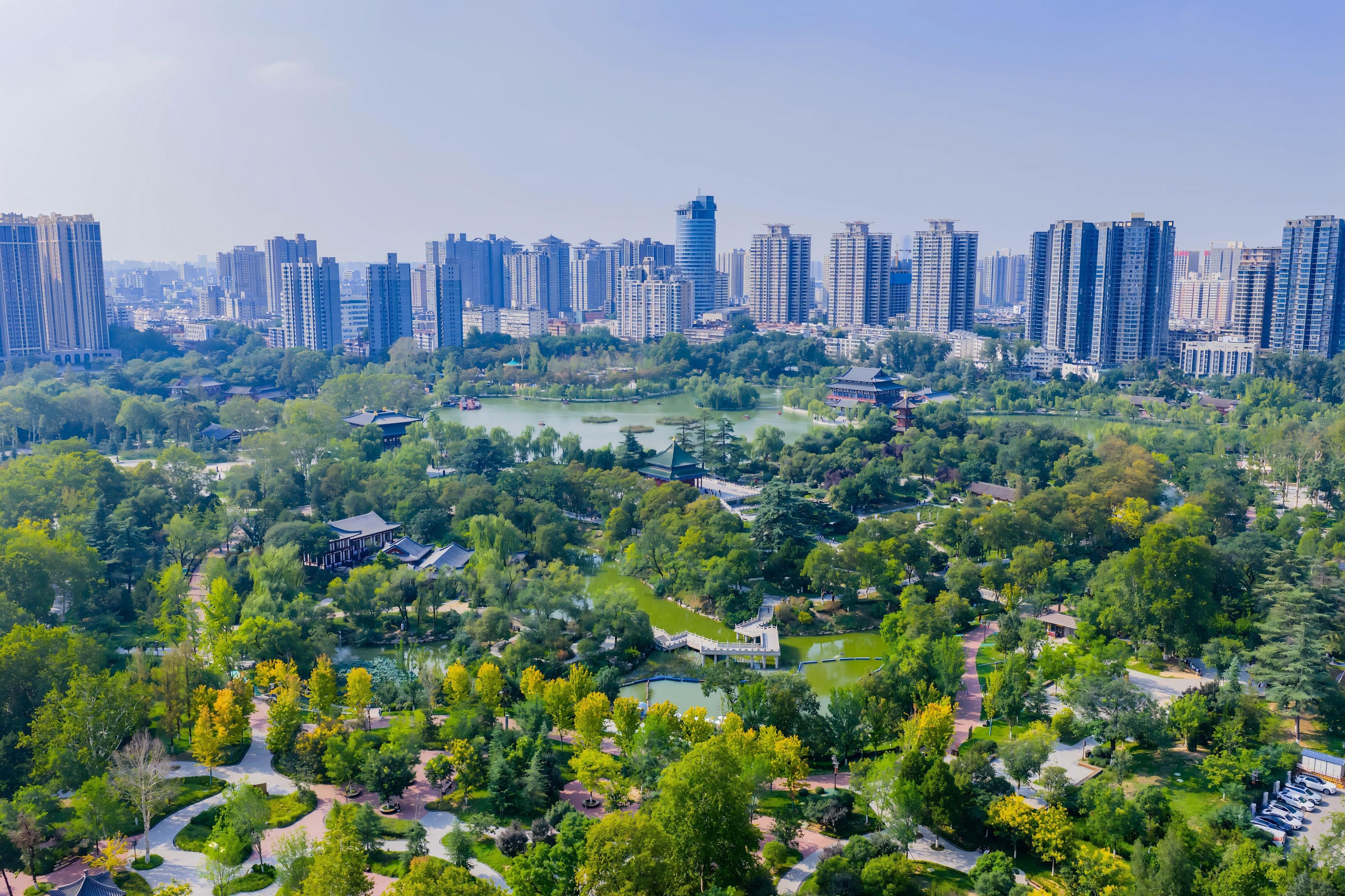 Um parque com caminhos para pedestres, um lago e arquitetura tradicional, integrado a uma paisagem urbana com edifícios altos.