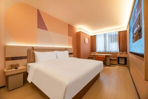 Quarto de hotel com uma cama grande, uma escrivaninha, uma cadeira, um pequeno frigorífico e uma televisão na parede.