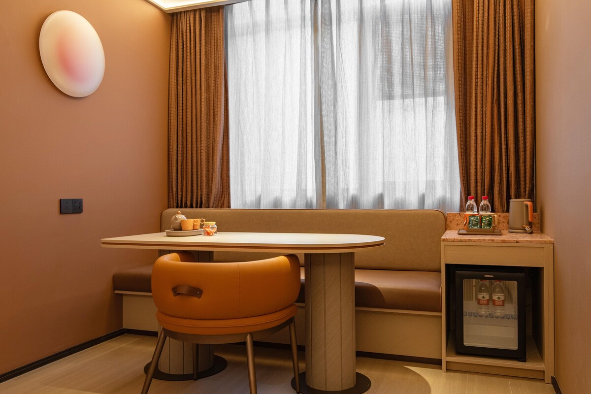 Quarto compacto com uma mesa de jantar, um sofá, um pequeno frigorífico e uma janela com cortinas.