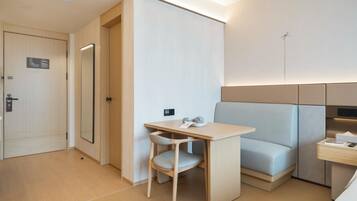 Quarto de hotel moderno com porta de madeira, uma pequena mesa com cadeira, cama com cabeceira e mesa de cabeceira com abajur.