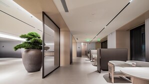 Um saguão de hotel moderno com uma grande planta em vaso, áreas de estar e um corredor longo.