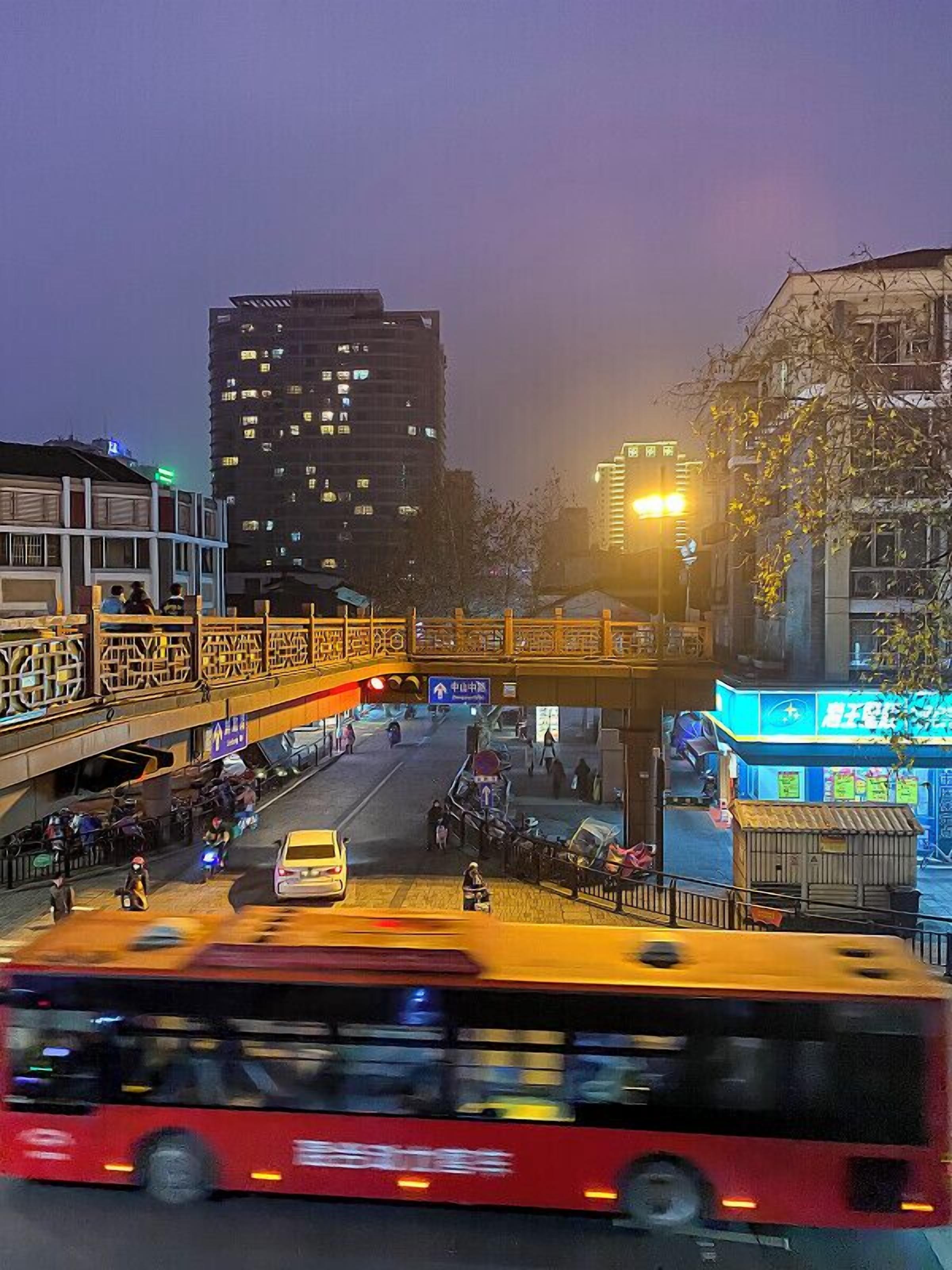 Rua movimentada em área urbana à noite, com um ônibus vermelho, uma passarela para pedestres e edifícios iluminados.