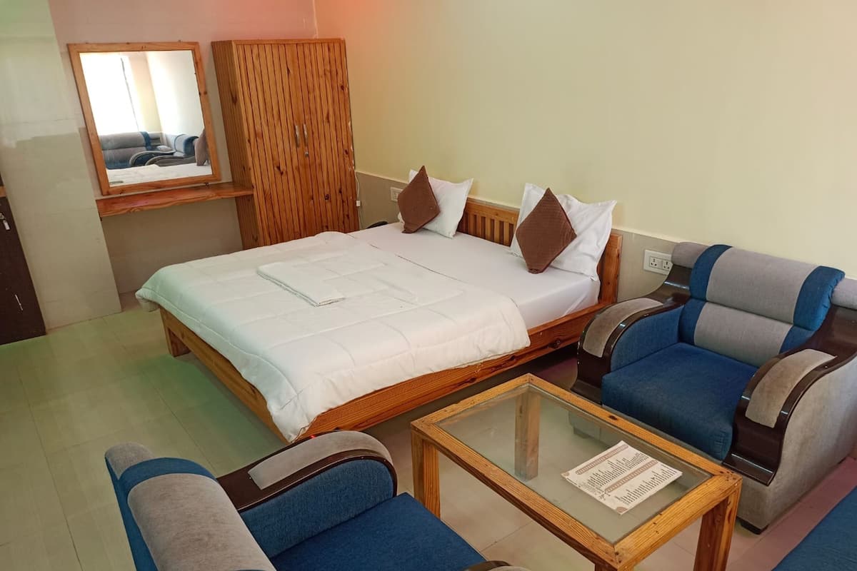 Quarto de hotel com cama, duas poltronas, uma mesa pequena, um guarda-roupa de madeira e um espelho.