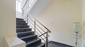 Uma escadaria com degraus de mármore preto e corrimãos de metal, um bebedouro à direita e uma parede em tom claro.