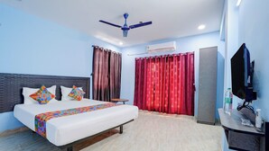 Quarto de hotel com cama, ventoinha no teto, ar condicionado, televisão e uma garrafa de água sobre uma mesa.