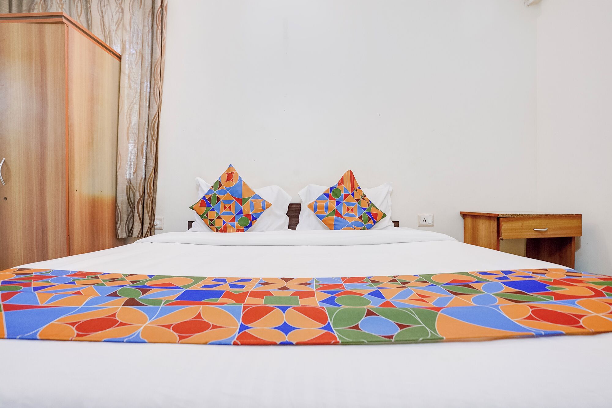 Uma cama com um edredom estampado em cores e padrões geométricos, duas almofadas com desenho similar, um guarda-roupa de madeira e uma pequena mesa de cabeceira de madeira.