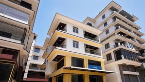 Um edifício residencial de vários andares com varandas, uma entrada visível e uma placa azul indicando um negócio ou serviço no andar térreo.