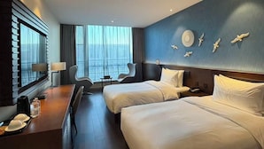Quarto de hotel com duas camas, uma escrivaninha, uma cadeira, televisão e vista para o mar.