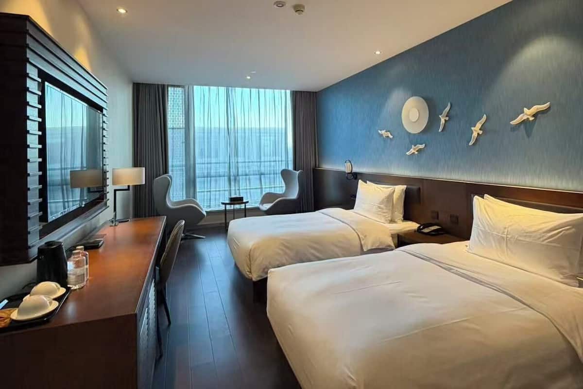 Quarto de hotel com duas camas, uma escrivaninha, uma cadeira, televisão e vista para o mar.