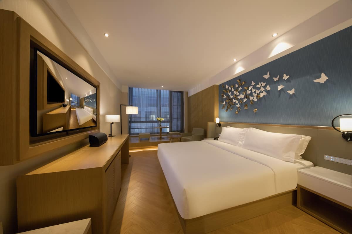 Quarto de hotel com uma cama grande, uma escrivaninha com computador, um espelho, uma parede decorada com borboletas e vista para a cidade à noite.