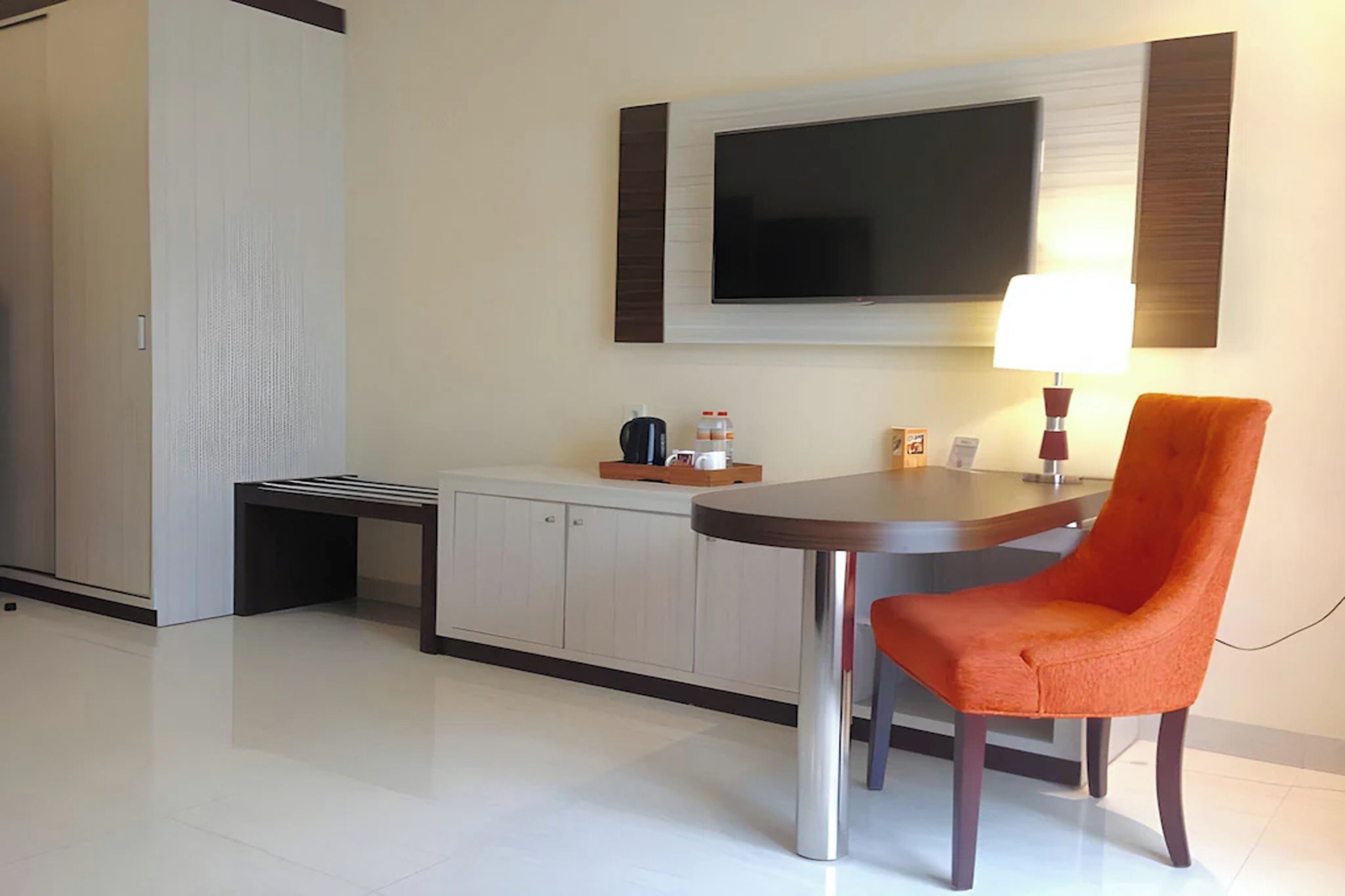 Quarto de hotel com uma mesa de jantar, uma única cadeira laranja, uma televisão fixada na parede e um pequeno armário com uma cafeteira.