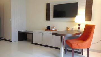 Quarto de hotel com uma mesa de jantar, uma única cadeira laranja, uma televisão fixada na parede e um pequeno armário com uma cafeteira.