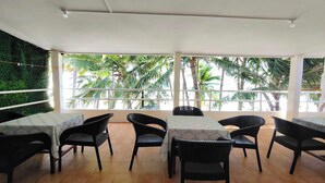 Restaurante com mesas brancas, cadeiras pretas e área externa, com vista para palmeiras.