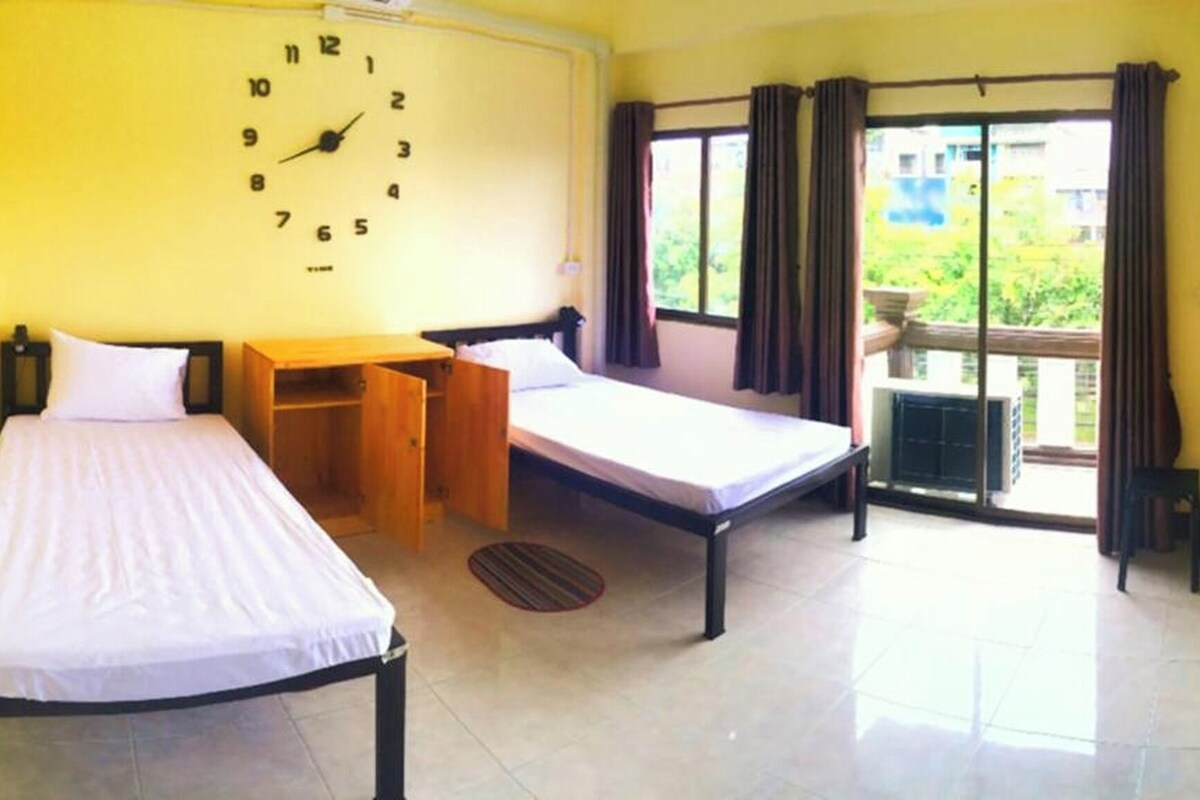Quarto com duas camas de solteiro, uma escrivaninha de madeira, um relógio de parede e uma porta de vidro deslizante que dá para uma área externa.