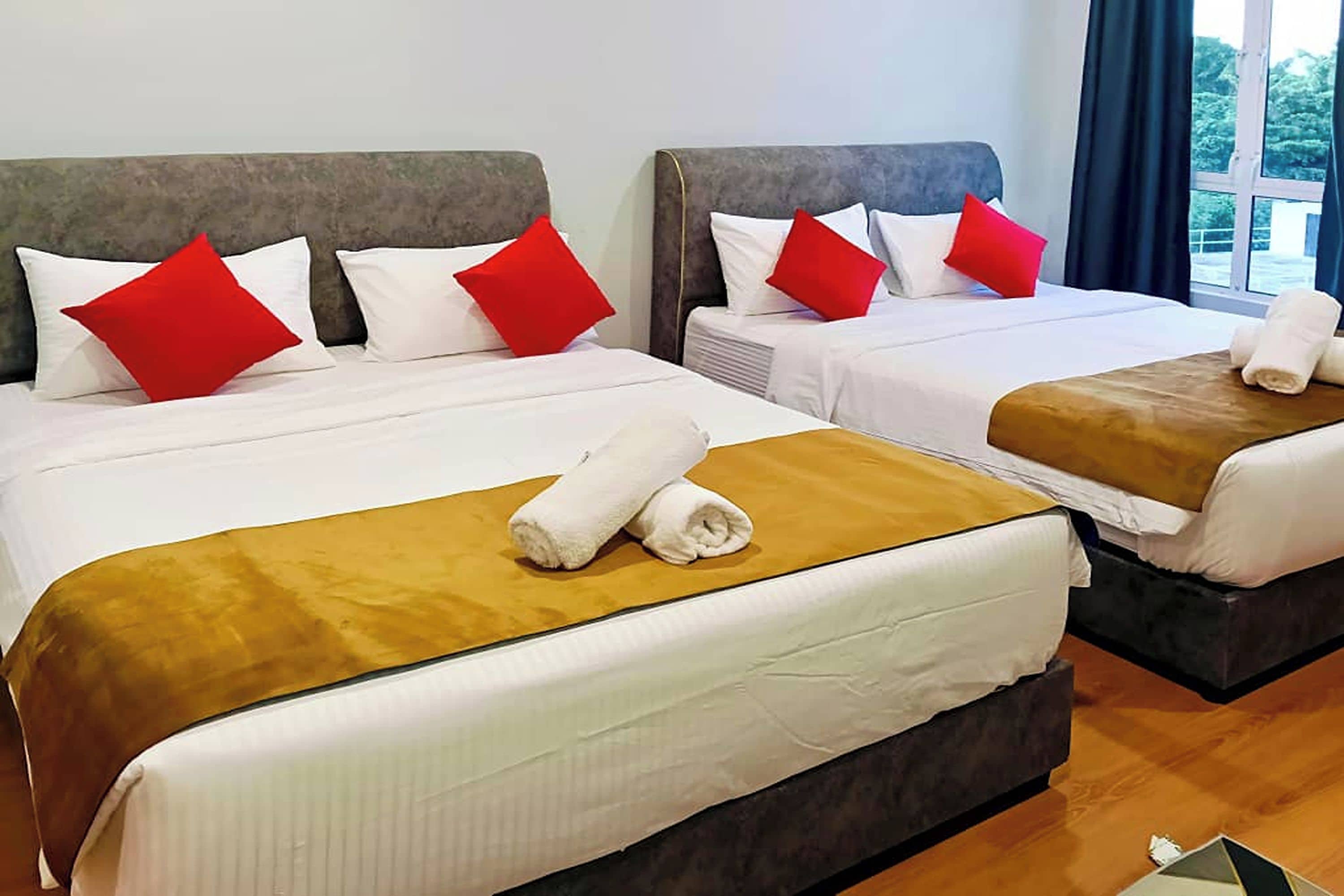 Quarto de hotel com duas camas, cada uma com roupa de cama branca e almofadas vermelhas, e cabeceira marrom.