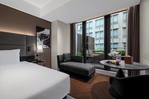 Quarto de hotel com cama, sofá, uma mesinha com frutas, luminária e vista para a cidade através de amplas janelas.