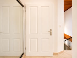 Uma porta branca fechada com puxador de latão, um interruptor de luz na parede e uma escada de madeira com corrimãos pretos.