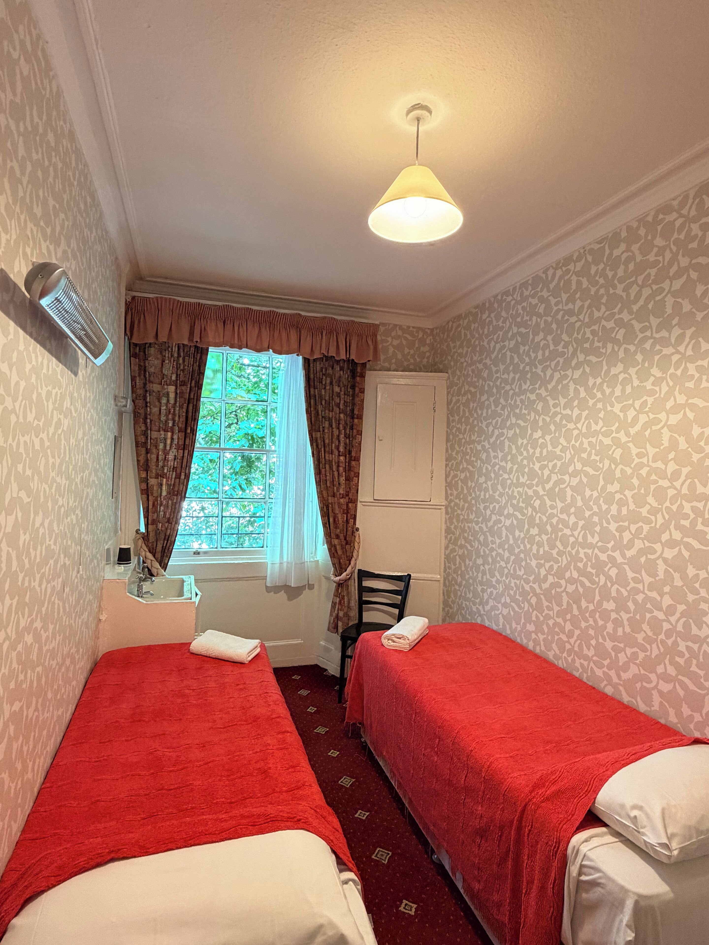 Quarto com duas camas, uma janela com cortinas, um radiador, uma pia e uma cadeira.
