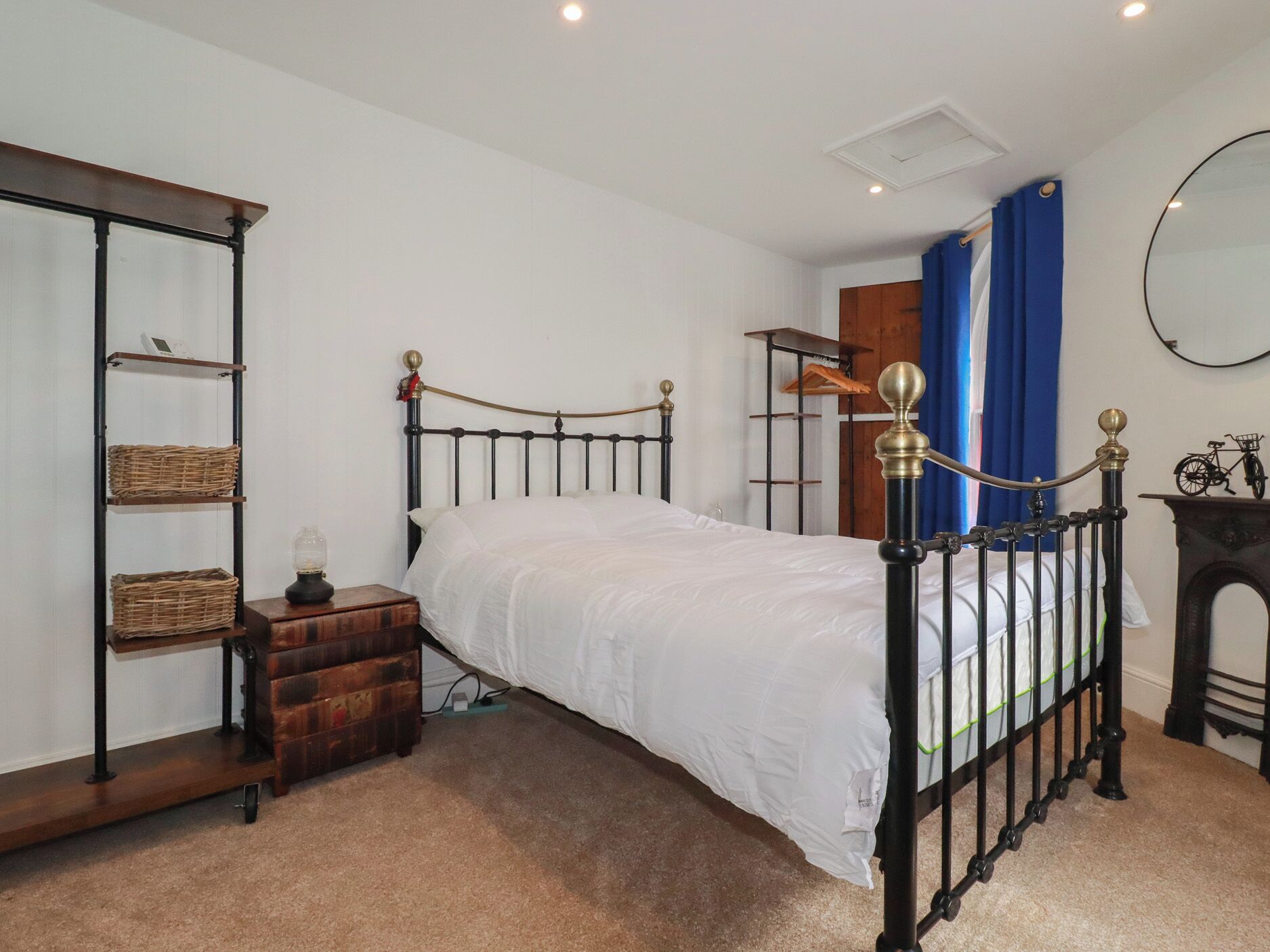 Um quarto com uma cama, uma mesinha de cabeceira de madeira, uma estante de vime, um espelho e uma cortina azul.