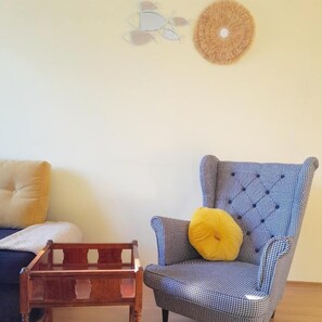 Um quarto com uma poltrona xadrez, uma pequena mesa de madeira, um travesseiro amarelo, um objeto decorativo na parede e um sofá com almofada amarela.