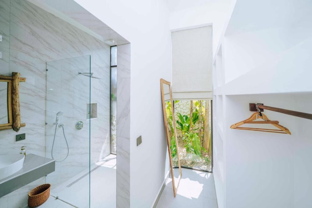 Casa de banho moderna com cabine de duche em vidro, lavatório branco e vista para o exterior arborizado.