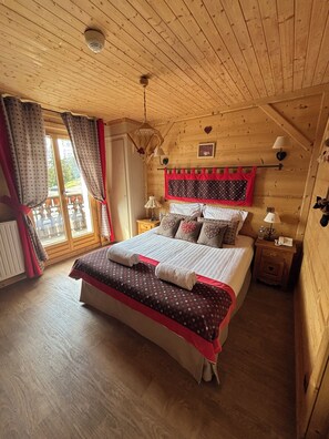 Quarto de cabana de madeira com cama, cortinas, janela, luminária fixada na parede e uma pequena mesa de cabeceira.