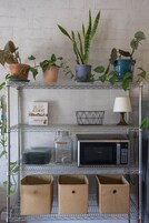 Uma estante de metal com plantas em vasos, um livro, um micro-ondas e cestos organizadores.