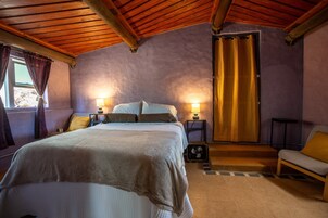 Quarto com uma cama, duas luminárias de cabeceira, uma cadeira, uma mesinha e uma janela com cortinas.