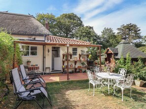 Uma casa com telhado de palha, um jardim com mesa e cadeiras de pátio, e diversos vasos de plantas.