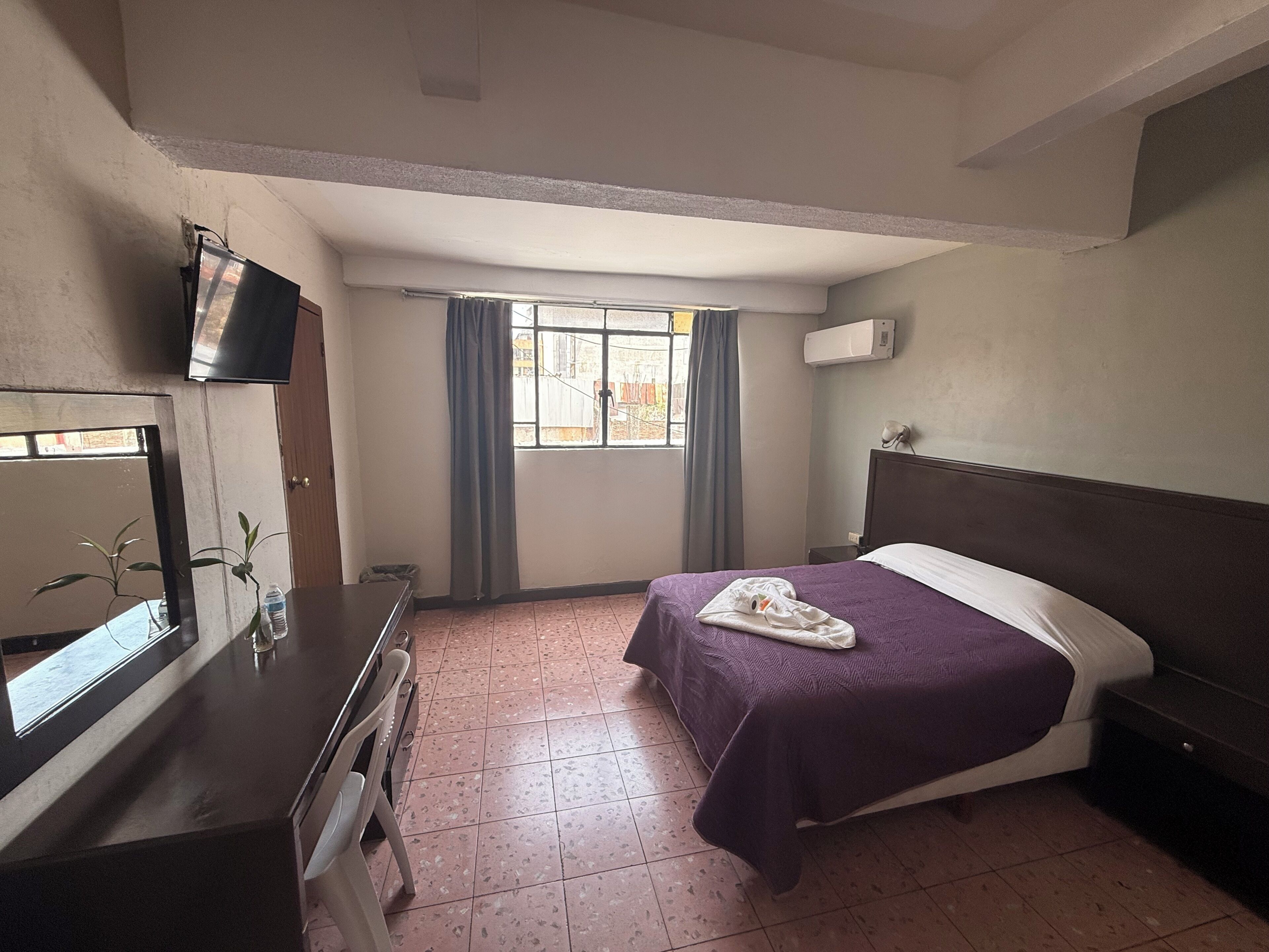 Quarto de hotel com uma cama, televisão em uma cômoda, janela com cortinas e piso ladrilhado.