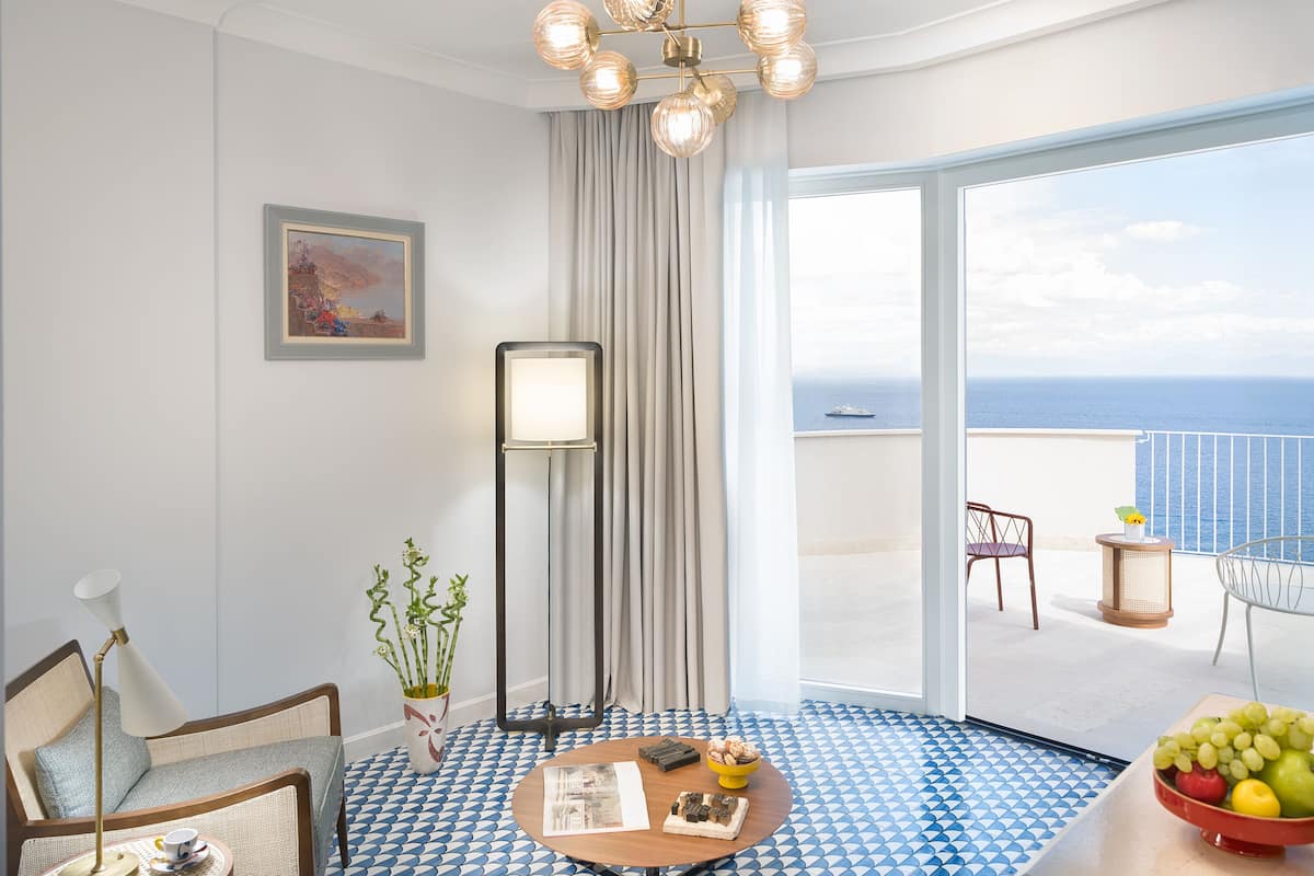 Quarto de hotel com varanda, uma mesa redonda com uma cesta de frutas, um abajur e uma pintura na parede.