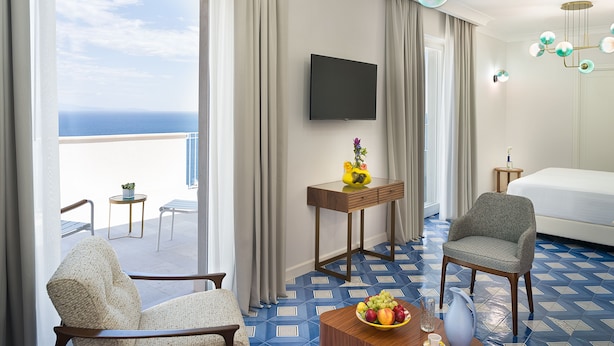 Quarto de hotel com uma grande janela, uma cama, uma TV, uma mesinha com uma cesta de frutas e uma cadeira.