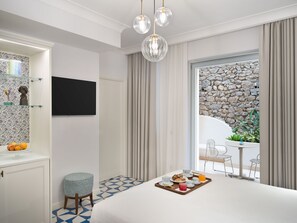 Quarto de hotel com cama, bandeja de café da manhã, televisão, um banquinho pequeno e vista para área externa com plantas.