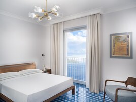 Quarto de hotel com cama, cadeira, quadro, janela com cortinas e luminária no teto.