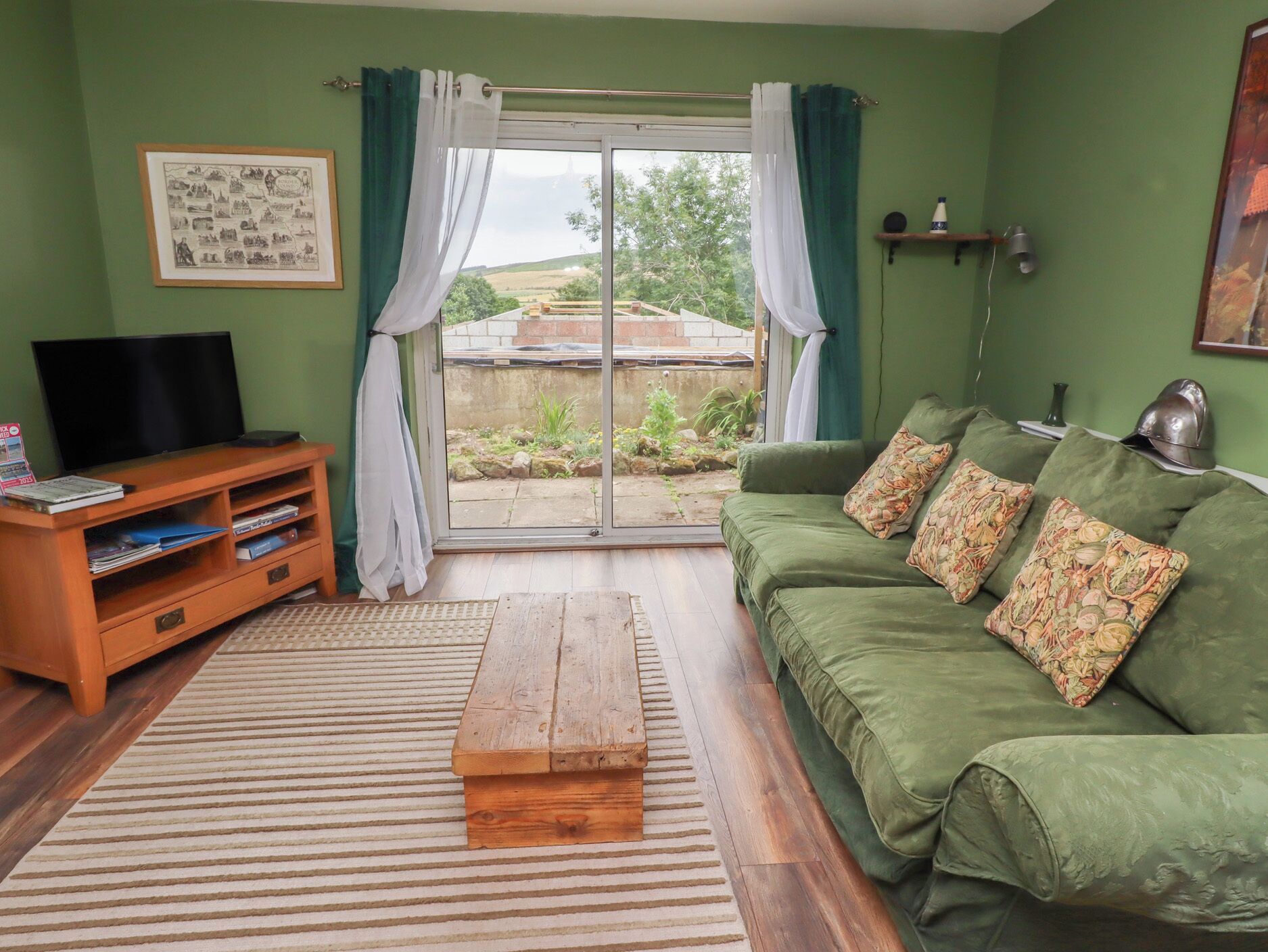 Uma sala de estar com um sofá verde, uma mesa de centro de madeira, um aparador com livros e uma janela grande com cortinas.