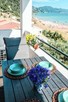 Varanda com mesa posta para dois, planta em vaso e vista para a praia e montanhas.