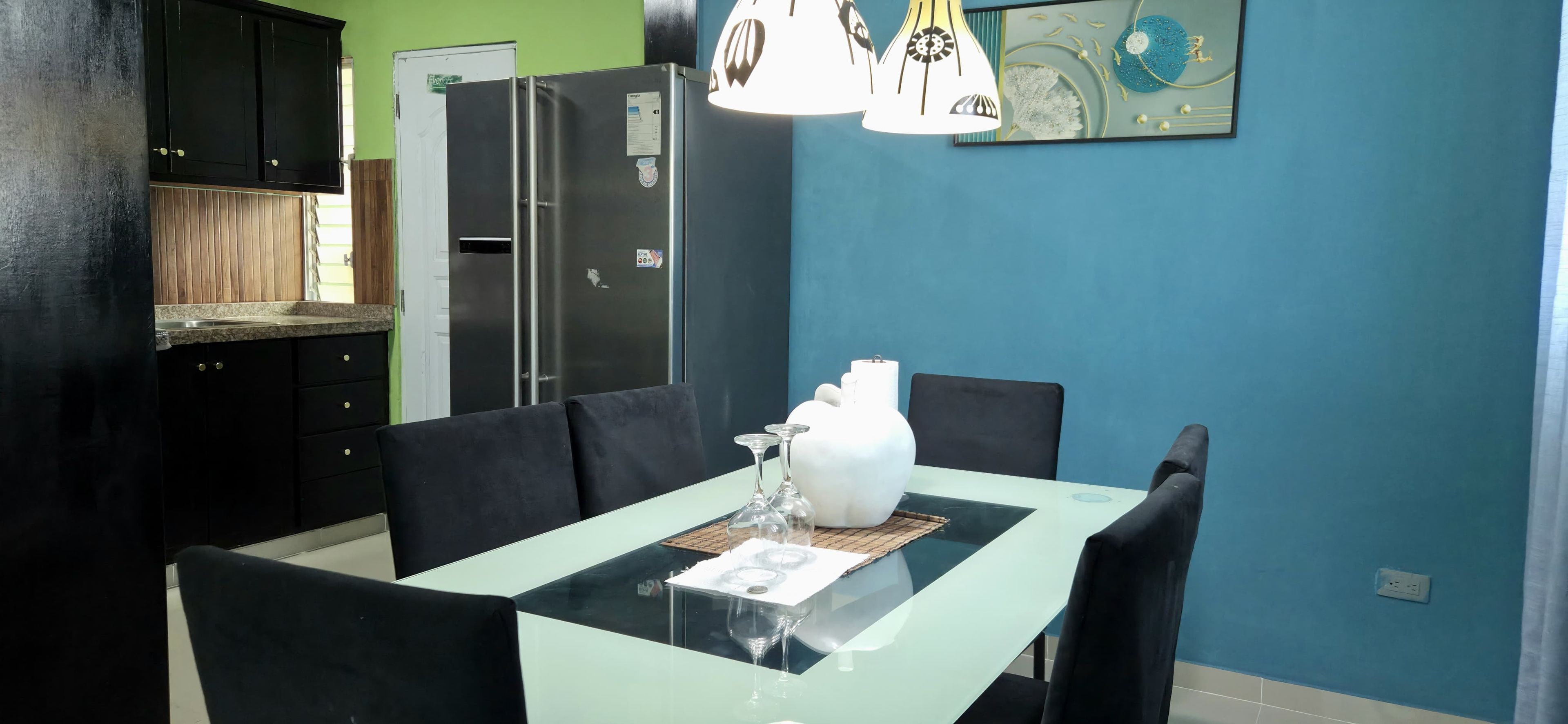 Uma sala de jantar com uma mesa posta para dois, geladeira e luminária fixada na parede.
