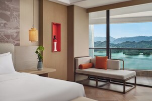 Quarto de hotel com uma cama, um sofá, um vaso com flores, uma estante vermelha e uma janela grande com vista para as montanhas.