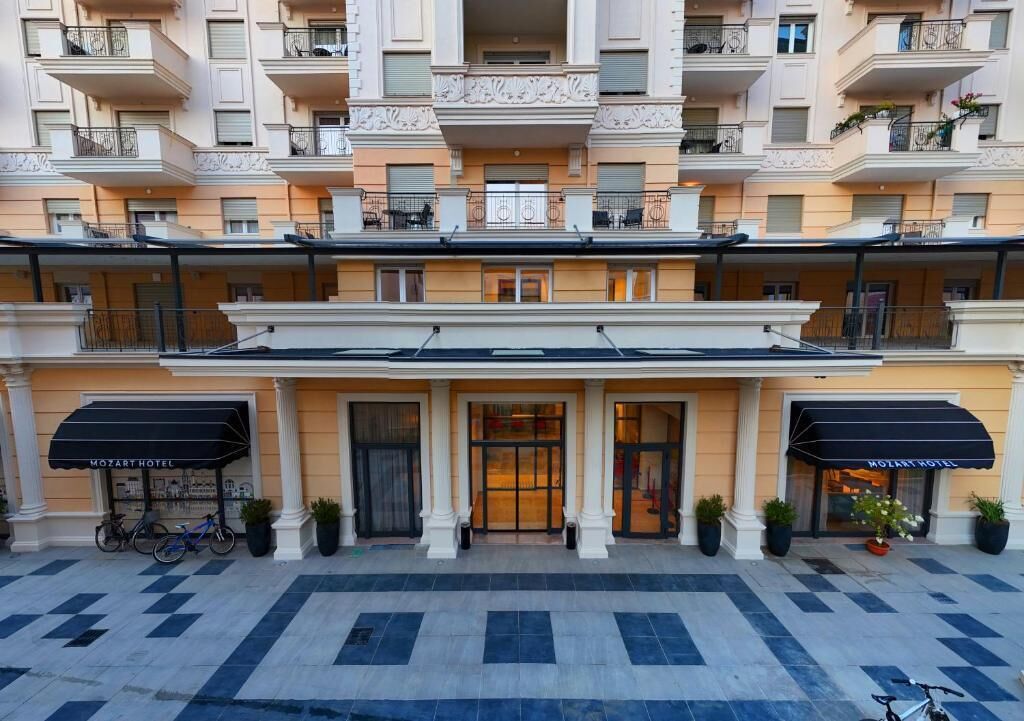 Um hotel com entrada central, varandas e uma praça calçada.