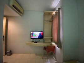Um quarto com televisão em um suporte, uma mesinha, uma cadeira, um cesto de lixo e uma porta.