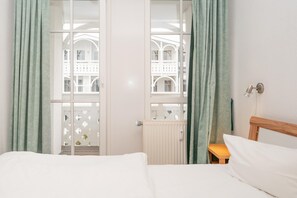 Quarto de hotel com cama, mesa de cabeceira, janela com cortinas e vista para um edifício com varandas.